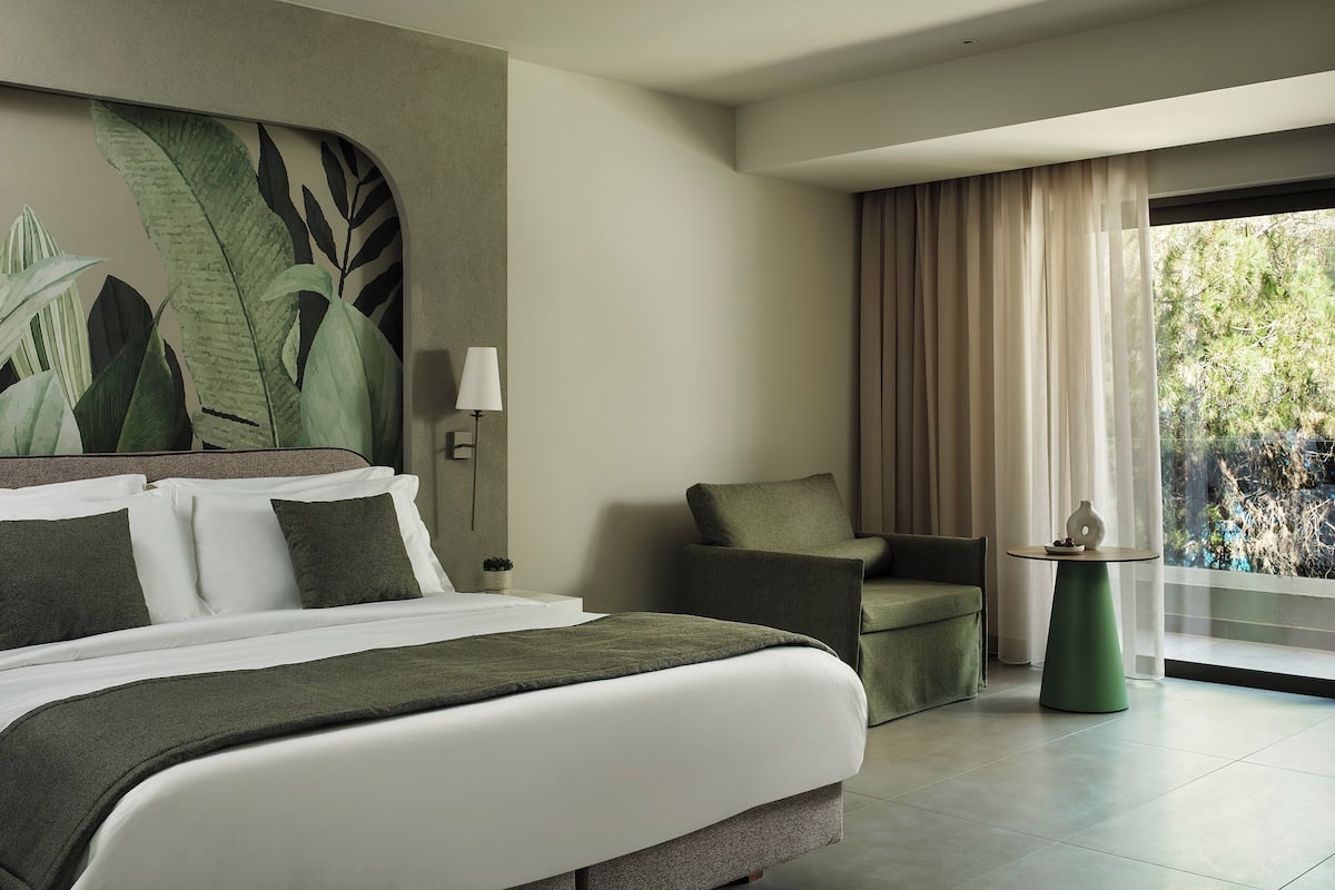 Quarto de hotel moderno com uma cama grande, uma poltrona verde, uma pequena mesa com um vaso e vista para o verde pela janela.