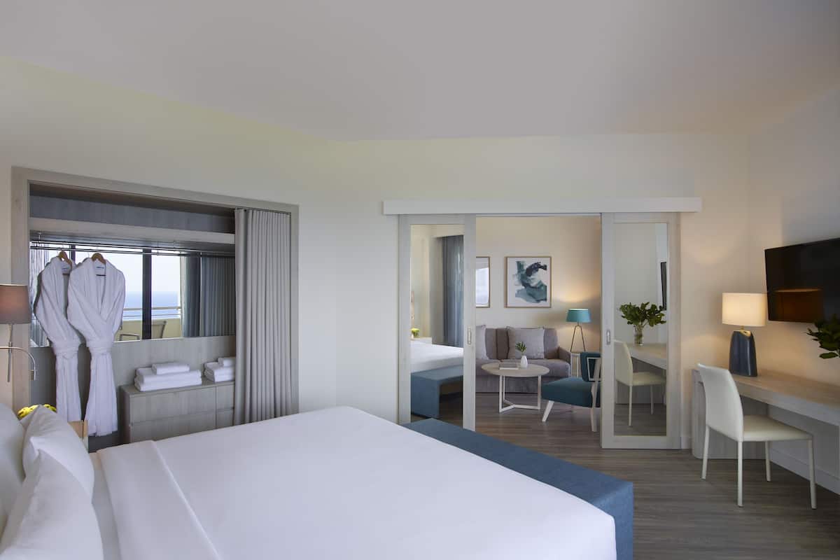 Quarto de hotel com uma cama grande, área de estar com sofá e poltronas, escrivaninha e televisão.