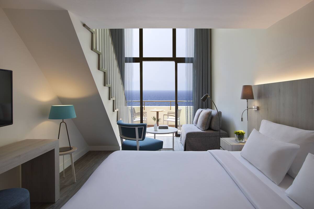 Quarto de hotel com uma cama grande, uma escrivaninha, uma cadeira, uma mesinha e vista para o oceano.