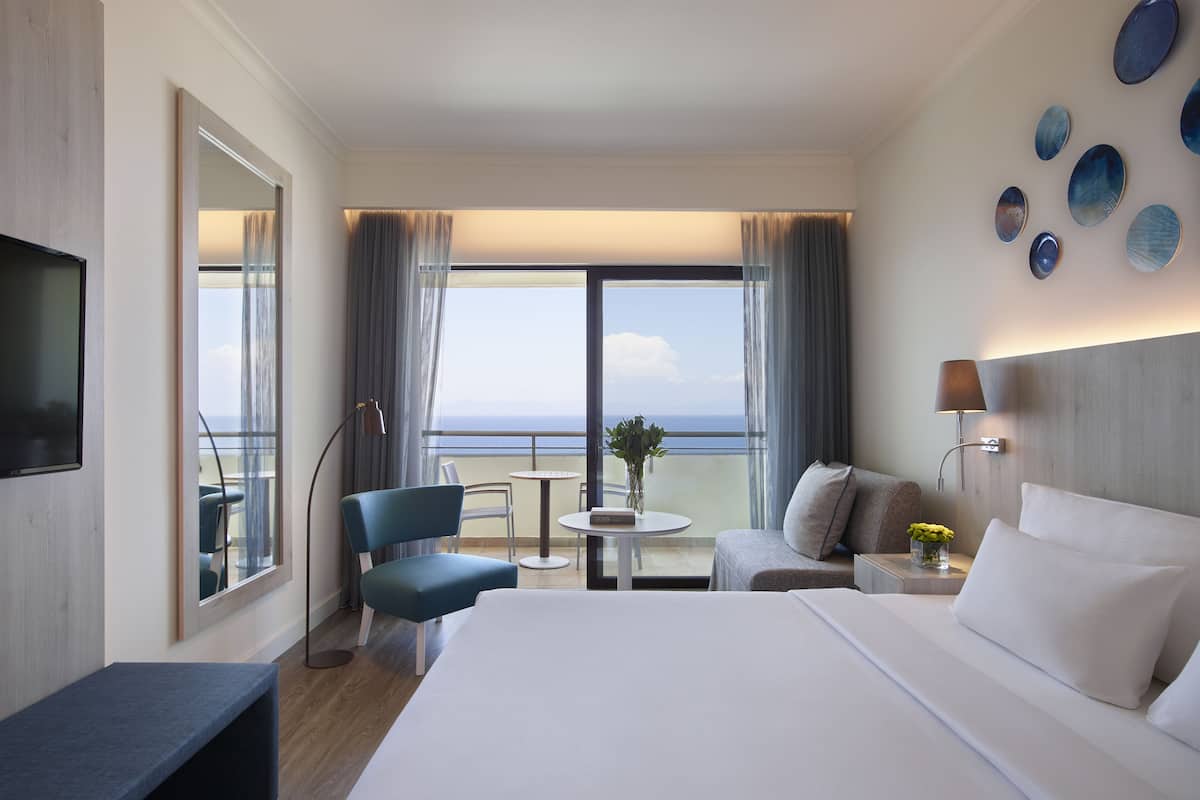 Quarto de hotel com uma cama grande, uma escrivaninha, uma cadeira, uma mesinha de centro com um vaso e vista para o mar.