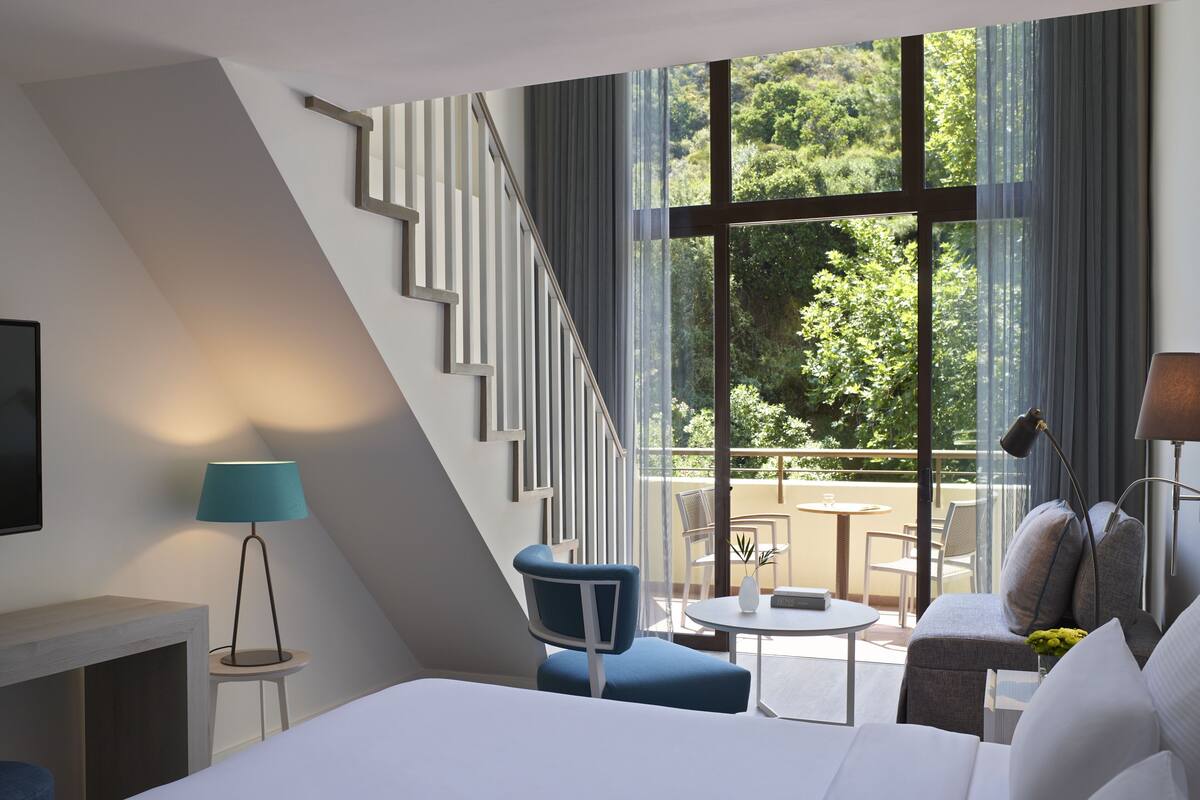 Quarto de hotel moderno com uma cama grande, uma mesa de escritório, uma cadeira, uma mesinha e vista para o exterior através de janelas amplas.