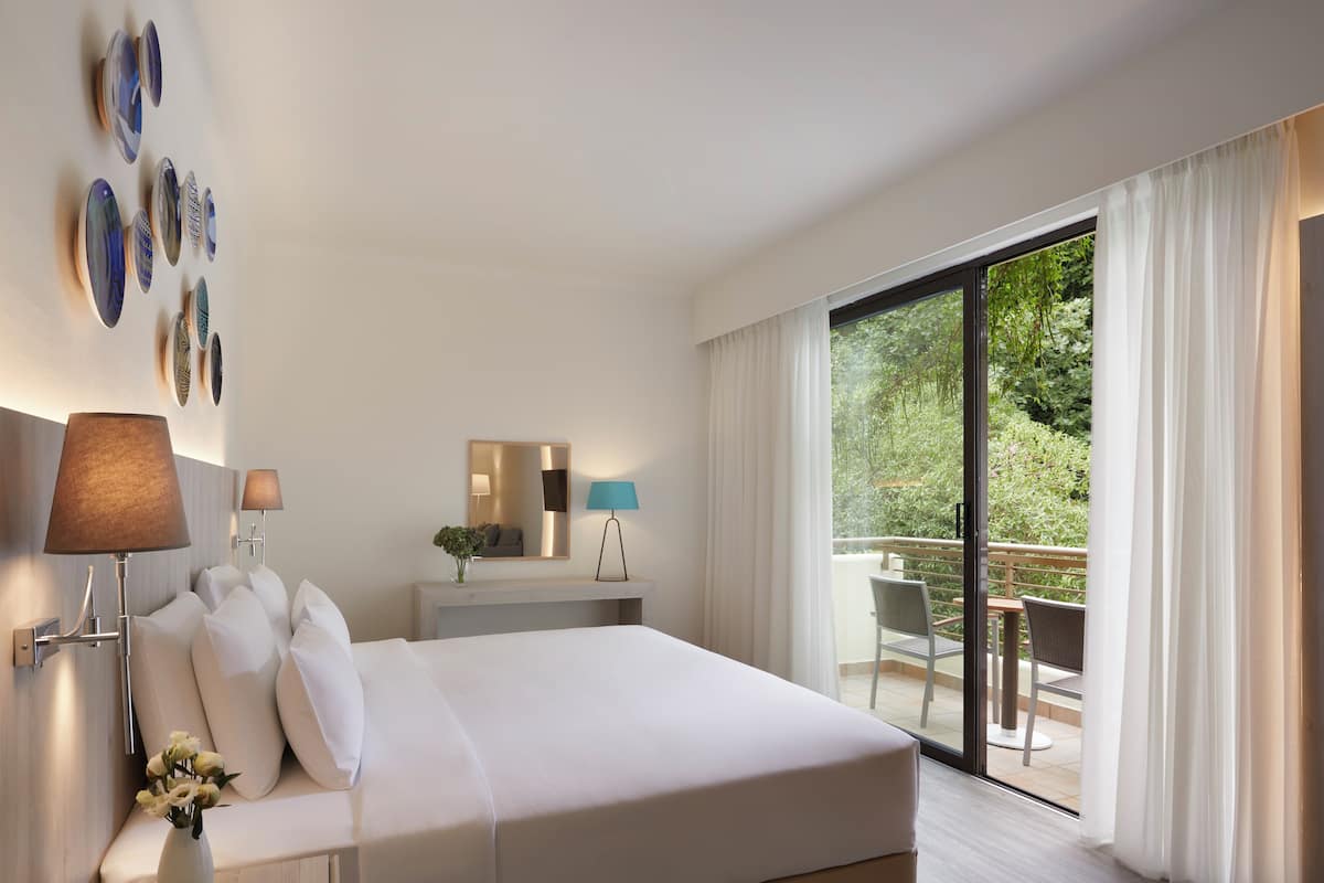 Quarto de hotel moderno com uma cama grande, mesa de cabeceira com luminárias, espelho e vista para uma varanda com mobiliário.