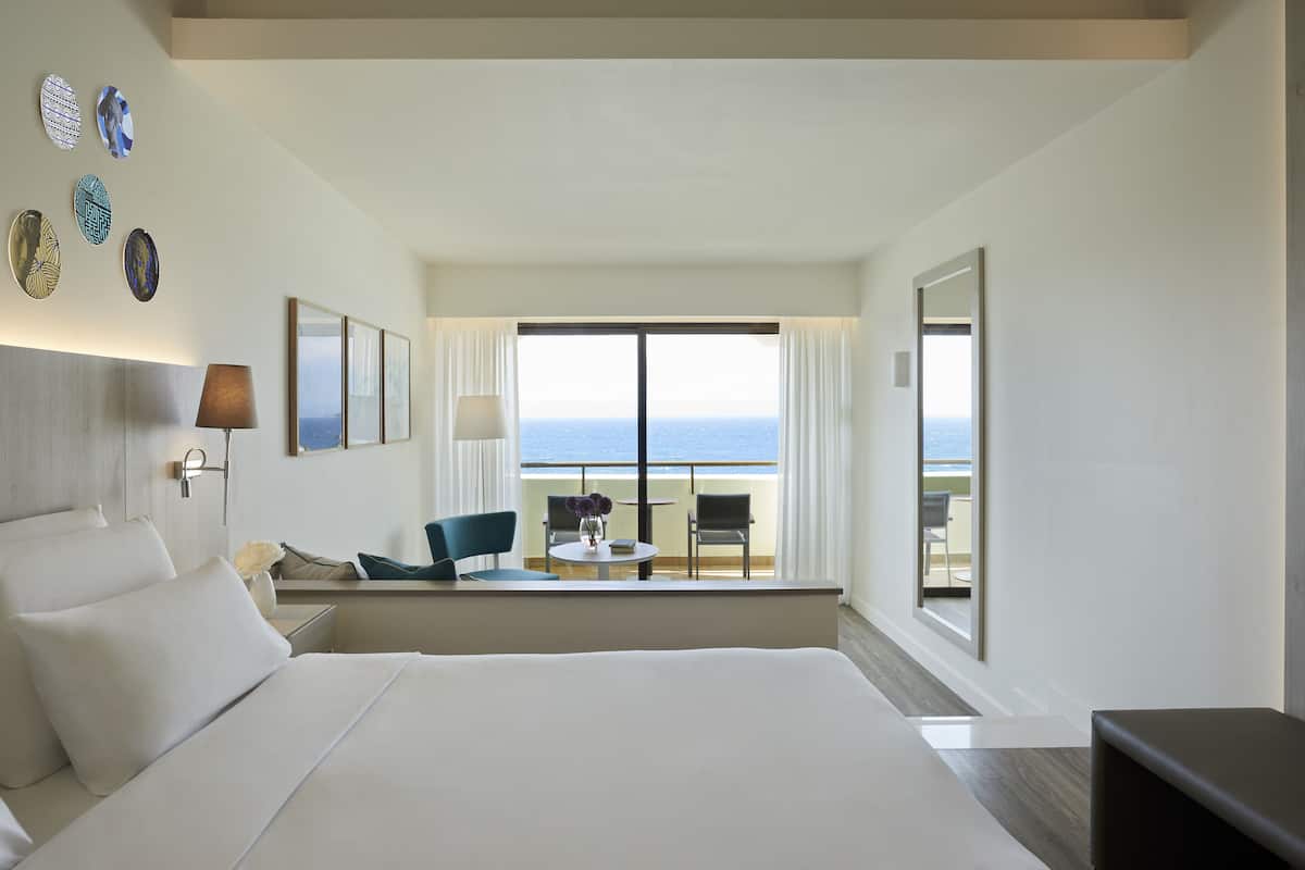 Quarto de hotel com uma cama grande, uma escrivaninha com cadeira, uma mesinha com duas cadeiras e varanda com vista para o oceano.