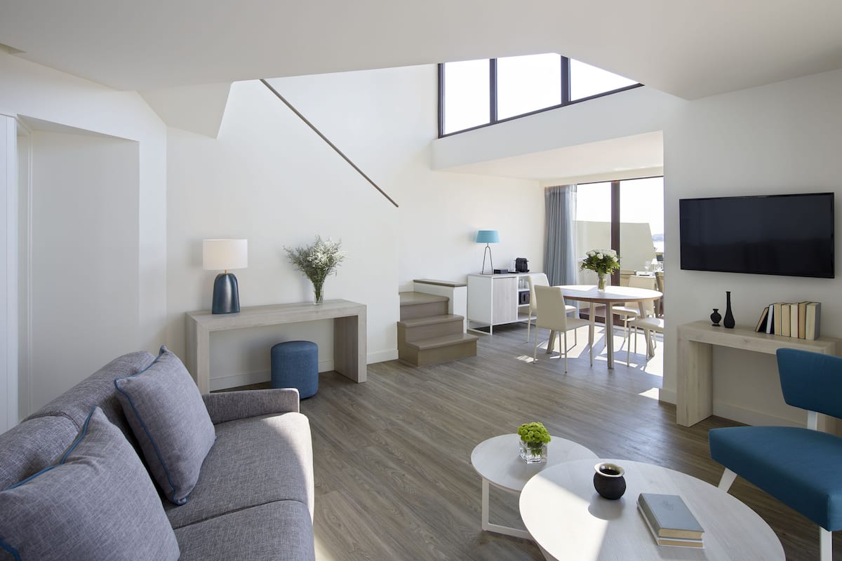 Uma sala de estar moderna com sofá, mesa de centro e televisão. Uma escadaria leva a uma área de jantar com mesa e cadeiras.