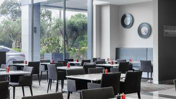 Um restaurante moderno com mesas, cadeiras, janelas amplas e vista para árvores e um carro do lado de fora.