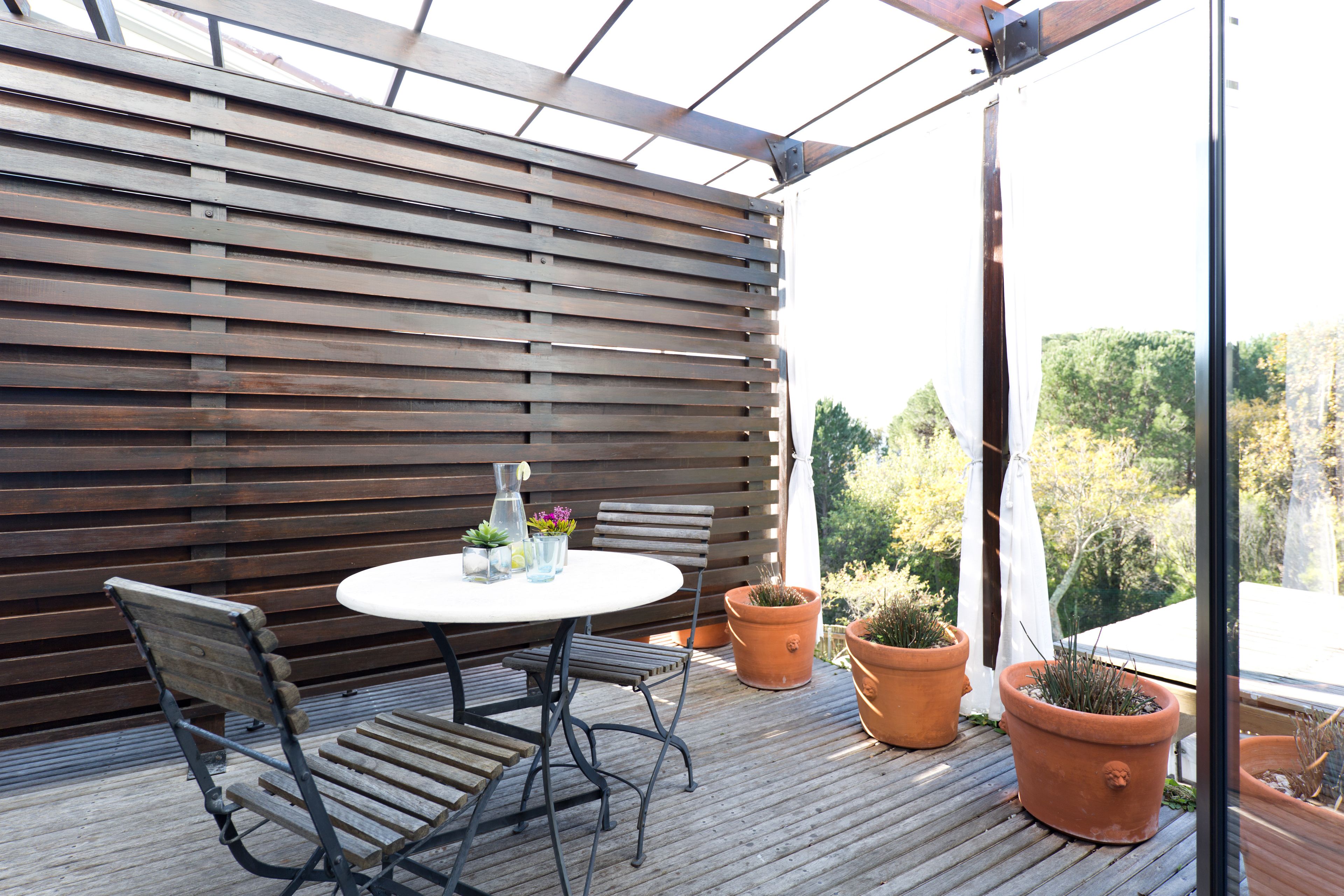 Um deck de madeira com mesa, cadeiras, plantas em vasos e vista para árvores através de uma porta de vidro.