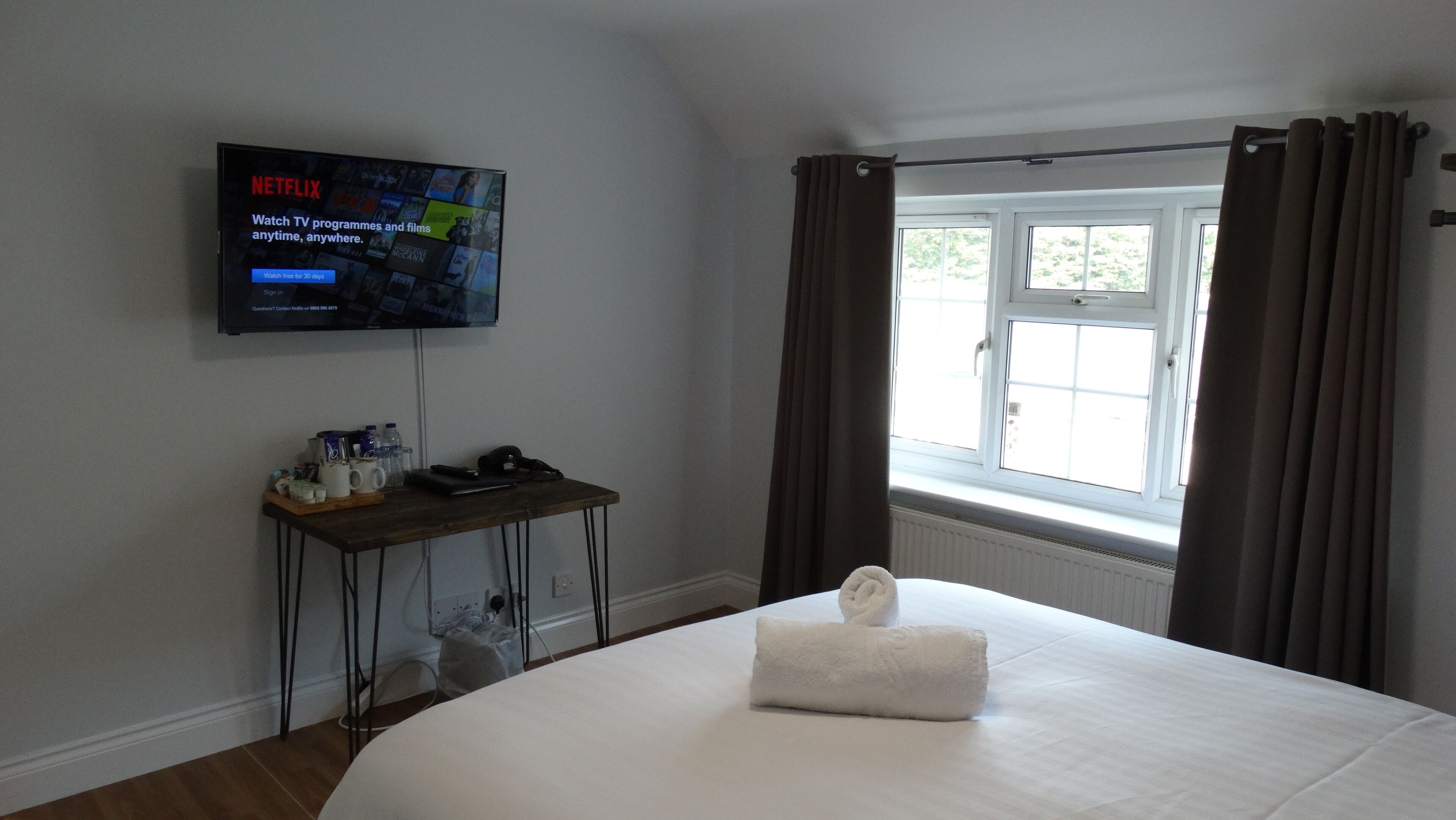 Quarto de hotel com uma cama, uma TV fixada na parede exibindo Netflix, uma pequena mesa com artigos de higiene e uma janela com cortinas.