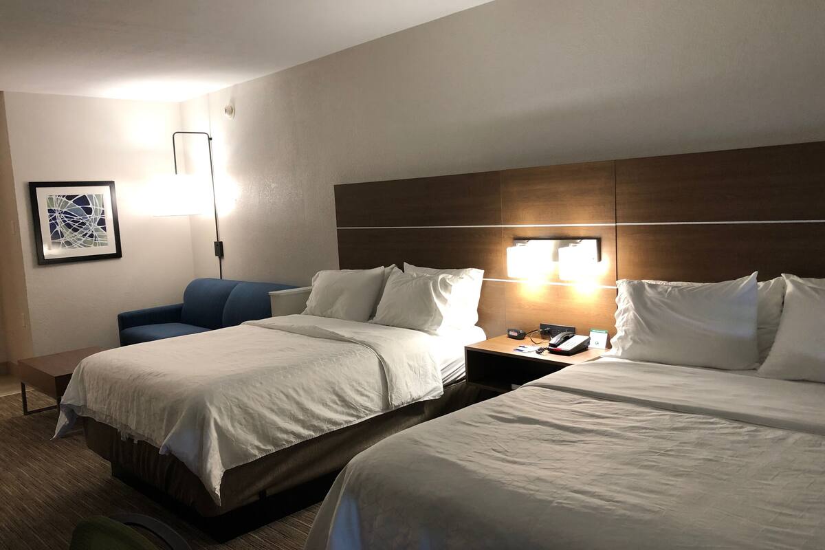 Quarto de hotel com duas camas, uma escrivaninha, uma cadeira, uma mesa de cabeceira com telefone e abajur, e um quadro na parede.