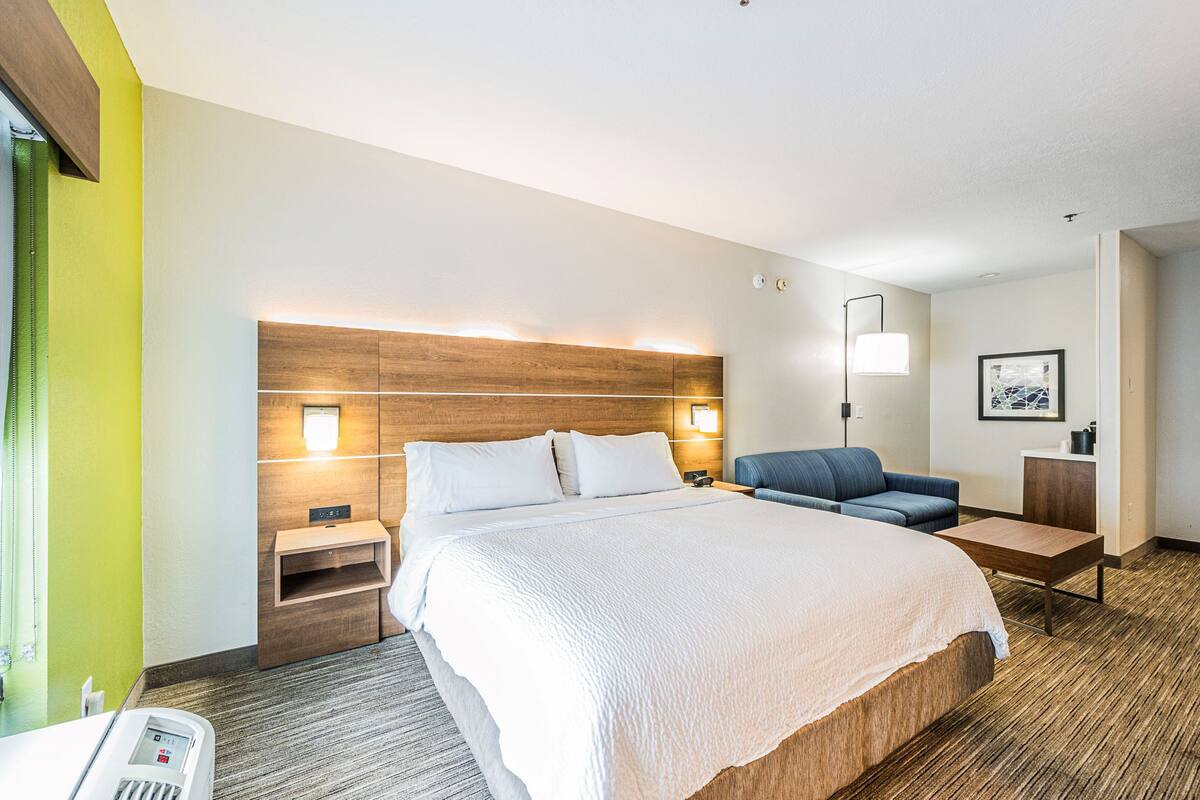 Quarto de hotel com uma cama grande, uma mesa de cabeceira, um criado-mudo, um sofá azul, uma pequena mesa de madeira e uma luminária na parede.