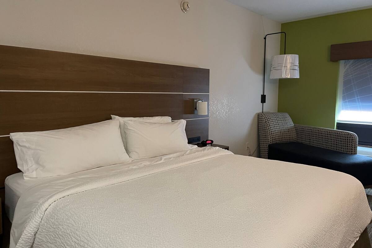 Cama bem arrumada com lençóis brancos, cabeceira de madeira, mesa de cabeceira com abajur e poltrona com padrão.