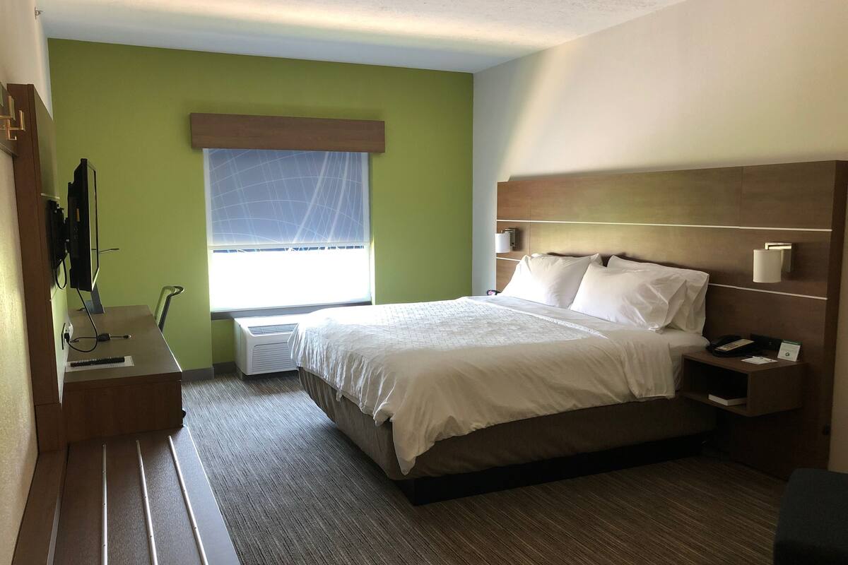 Quarto de hotel com cama, mesa de cabeceira, abajur e janela com persianas.