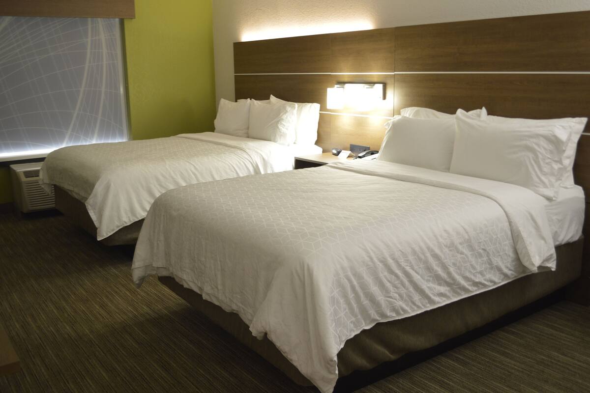 Quarto de hotel com duas camas, cabeceira de madeira, mesa de cabeceira com abajur e janela com persianas.