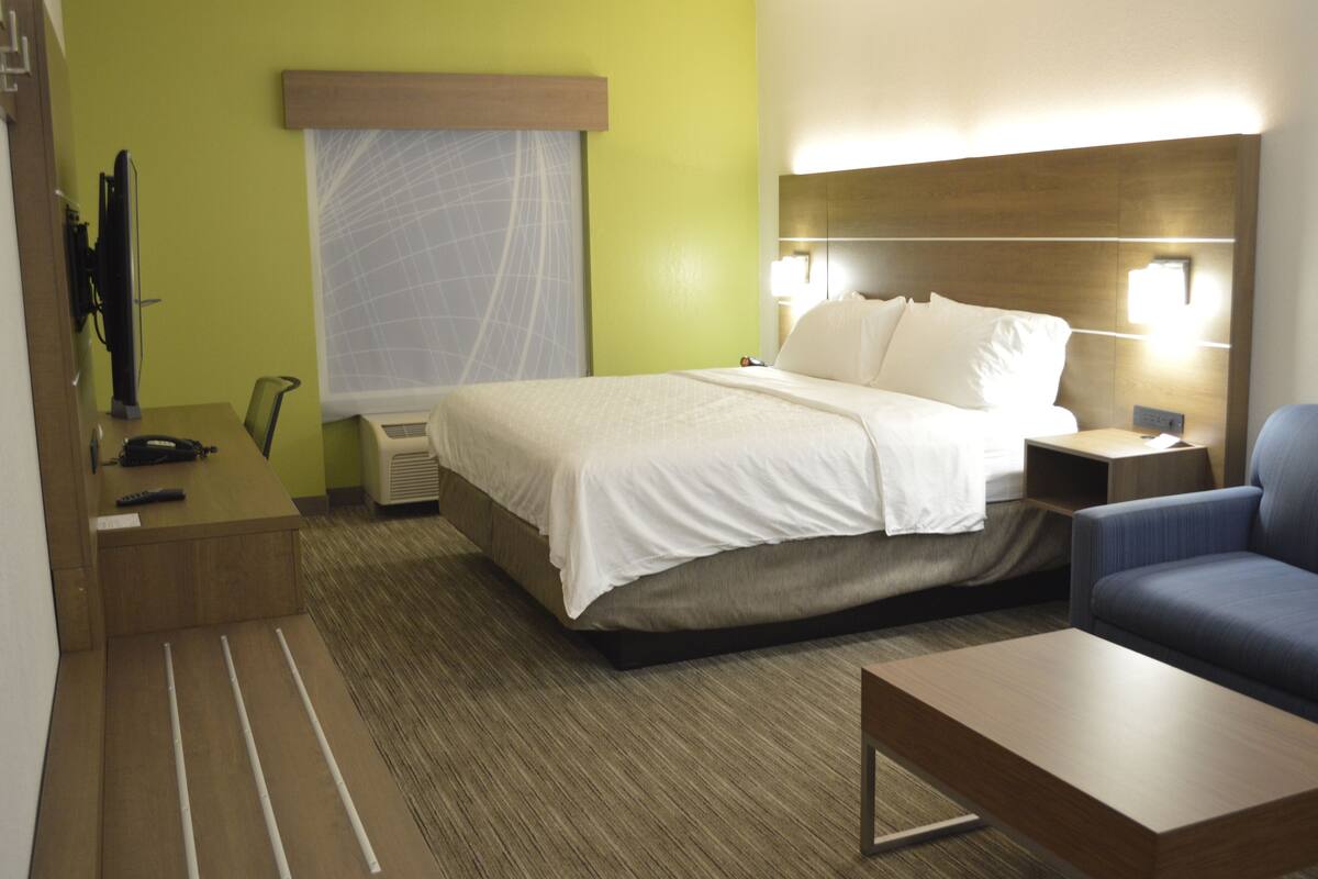 Quarto de hotel com cama, escrivaninha, cadeira, televisão e janela com persianas.
