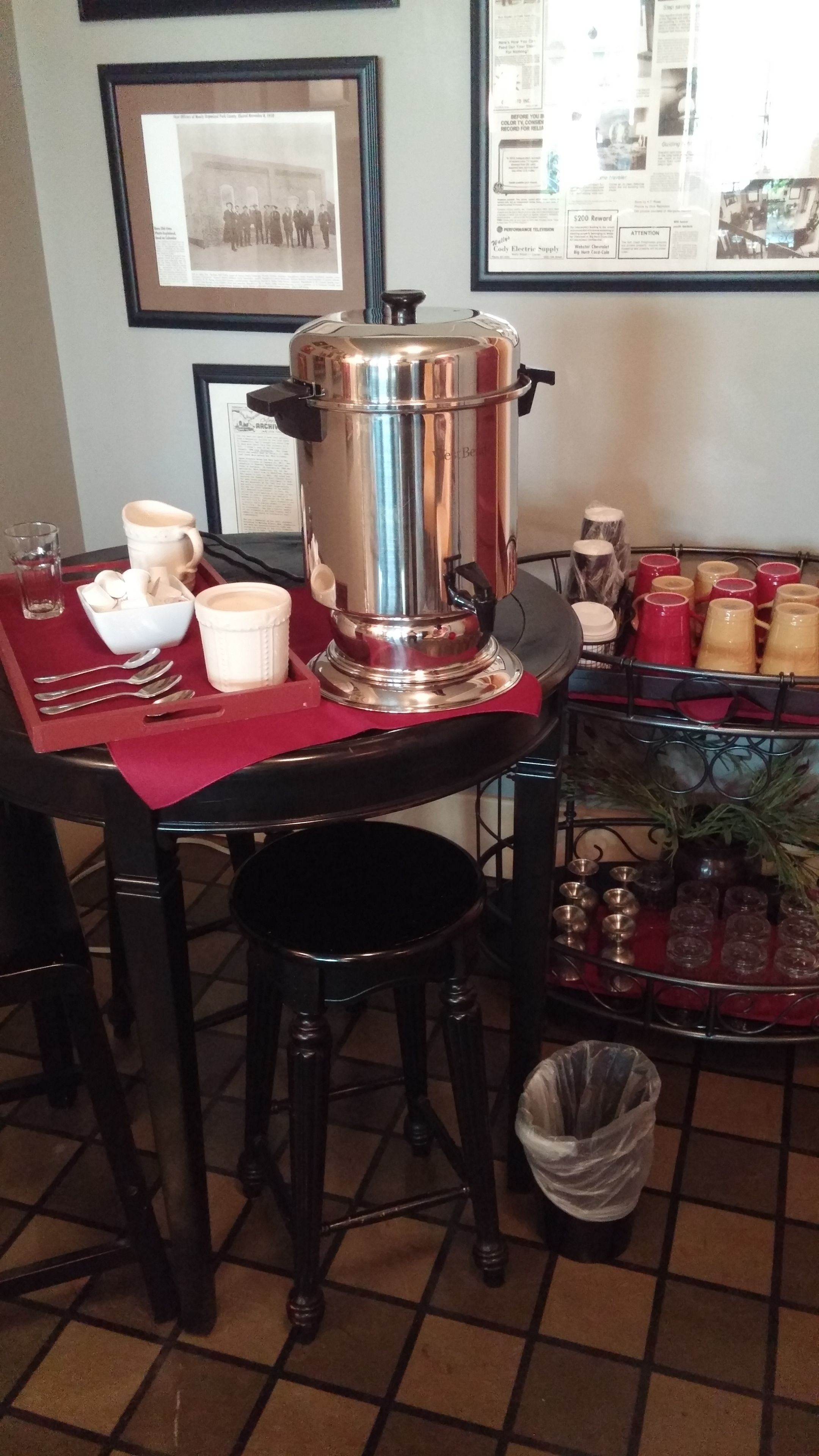 Uma área de cozinha com uma panela de aço inoxidável grande sobre uma mesa, toalha de mesa vermelha e diversos utensílios de cozinha, incluindo xícaras e uma tigela.