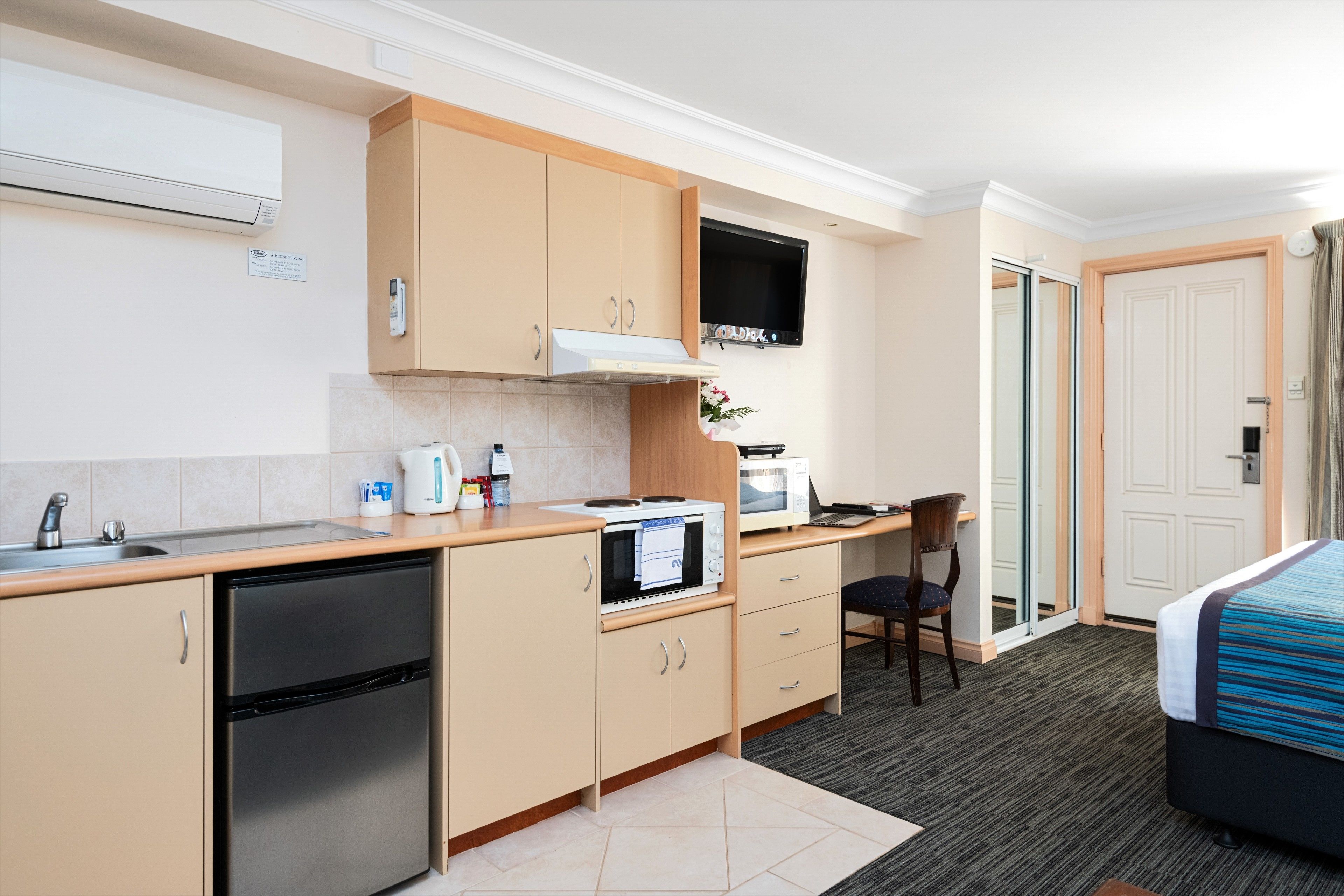 Quarto de hotel compacto com cozinha compacta, incluindo geladeira, micro-ondas e cafeteira. Há uma cama com manta listrada e uma cadeira.
