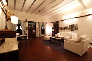 Uma sala de estar ampla com um sofá branco, uma mesa de centro de madeira e uma mesa de mármore. Há quadros nas paredes e um vaso com flores sobre uma mesa lateral.