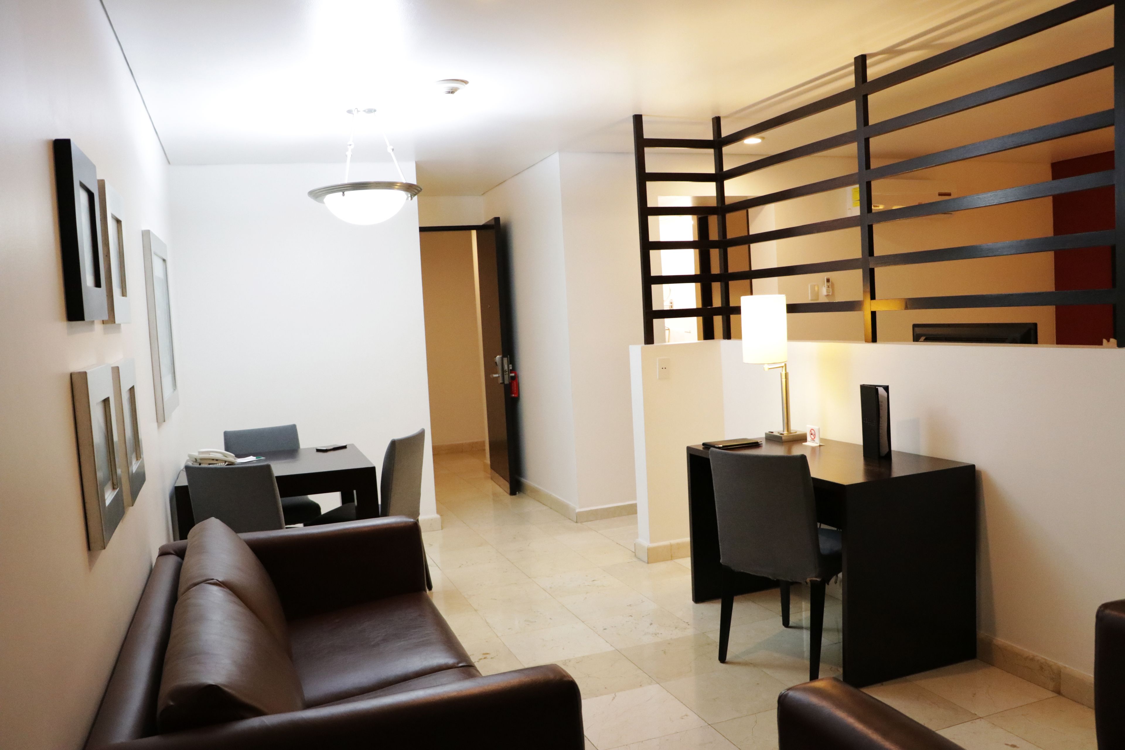 Uma sala de estar moderna com sofá, mesa de jantar e escrivaninha com abajur. Há uma divisória com ripas horizontais e uma porta que dá para outro cômodo.