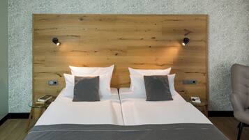 Quarto de hotel com cabeceira de madeira, duas camas com roupa de cama branca e cinza, mesas de cabeceira com abajures e uma cadeira.