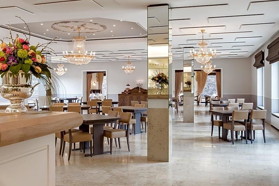 Ampla área de refeições de hotel, com diversas mesas e cadeiras, lustres e um espelho grande que reflete o espaço.