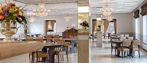 Ampla área de refeições de hotel, com diversas mesas e cadeiras, lustres e um espelho grande que reflete o espaço.