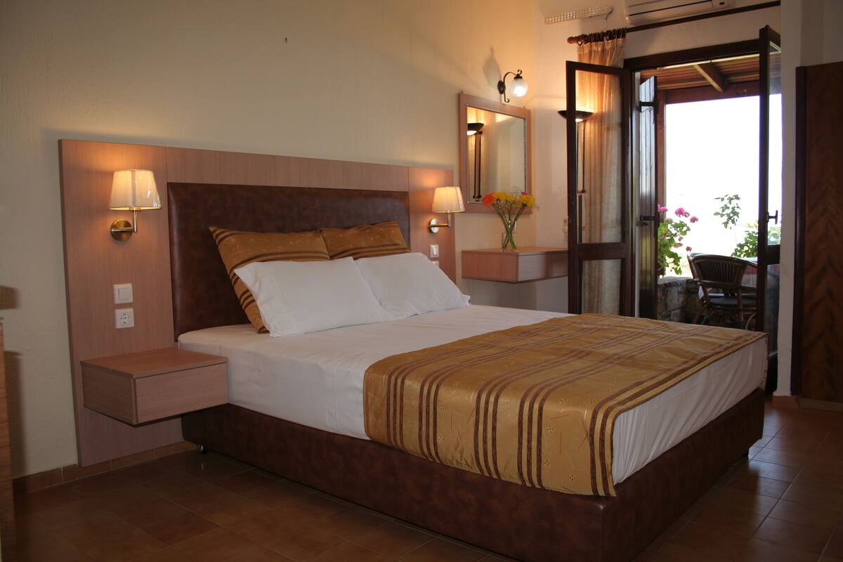 Quarto de hotel com uma cama grande, cabeceira de madeira, abajures, mesa de cabeceira e vista para o exterior através de uma porta de vidro.