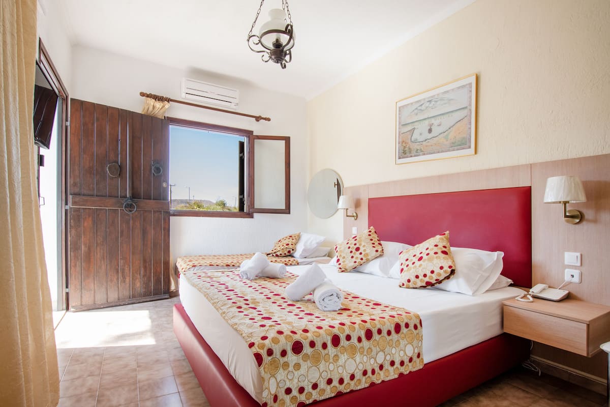 Quarto de hotel com uma cama grande, um guarda-roupa de madeira, mesa de cabeceira com abajur e vista para o exterior.