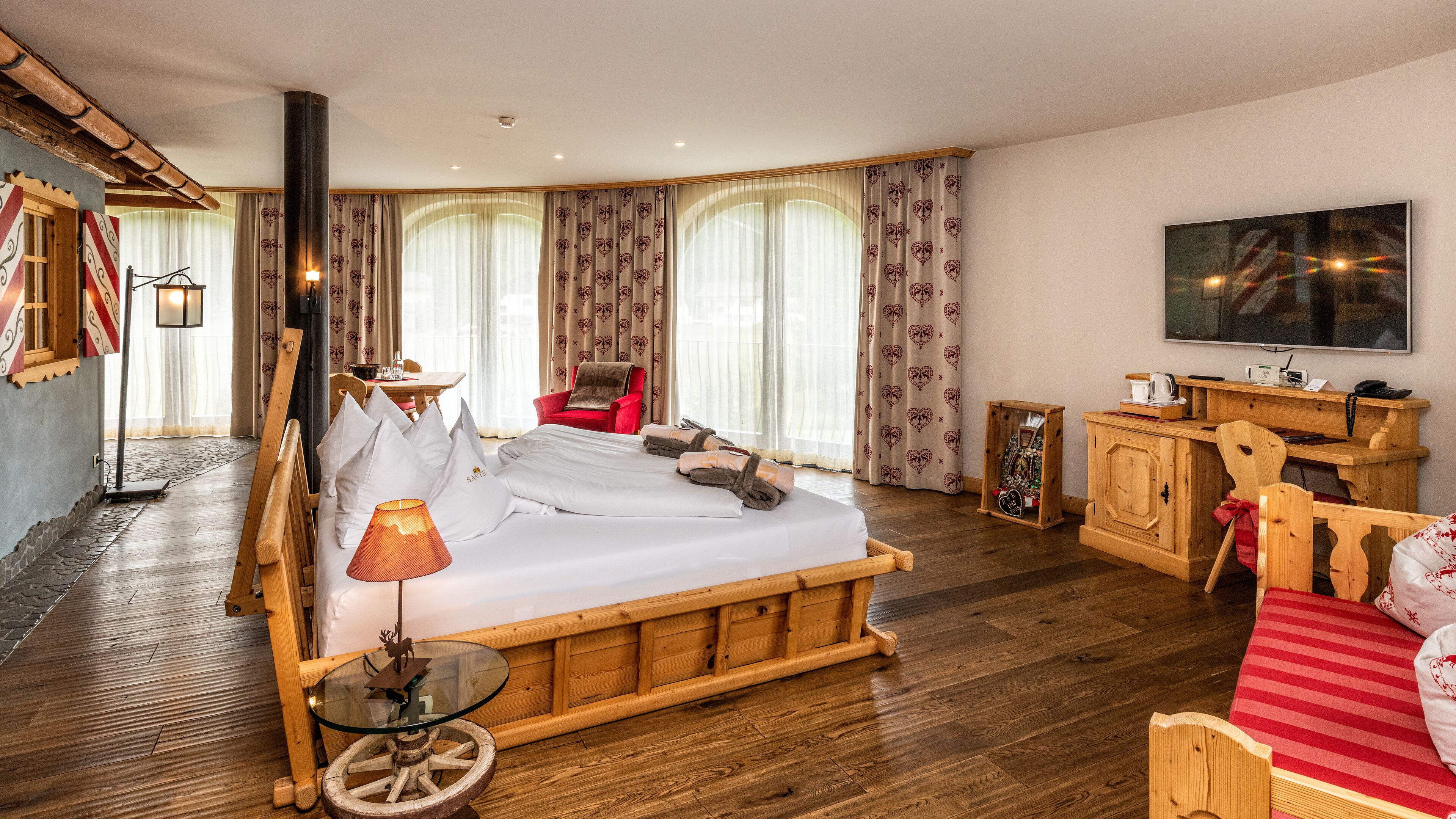 Quarto de hotel aconchegante com cama de madeira, mesa de vidro, cadeira vermelha, escrivaninha de madeira e televisão.