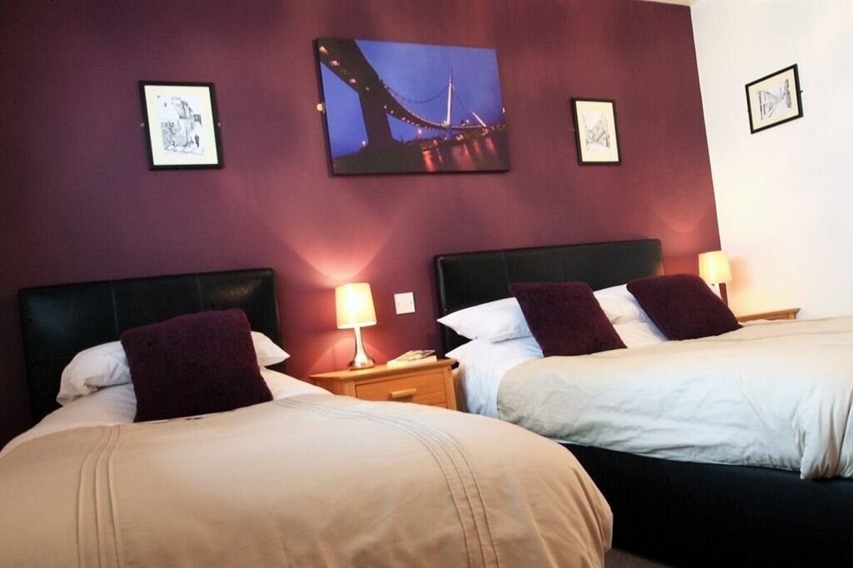 Quarto de hotel com duas camas, cada uma com cabeceira, mesa de cabeceira com abajur e quadro emoldurado na parede.