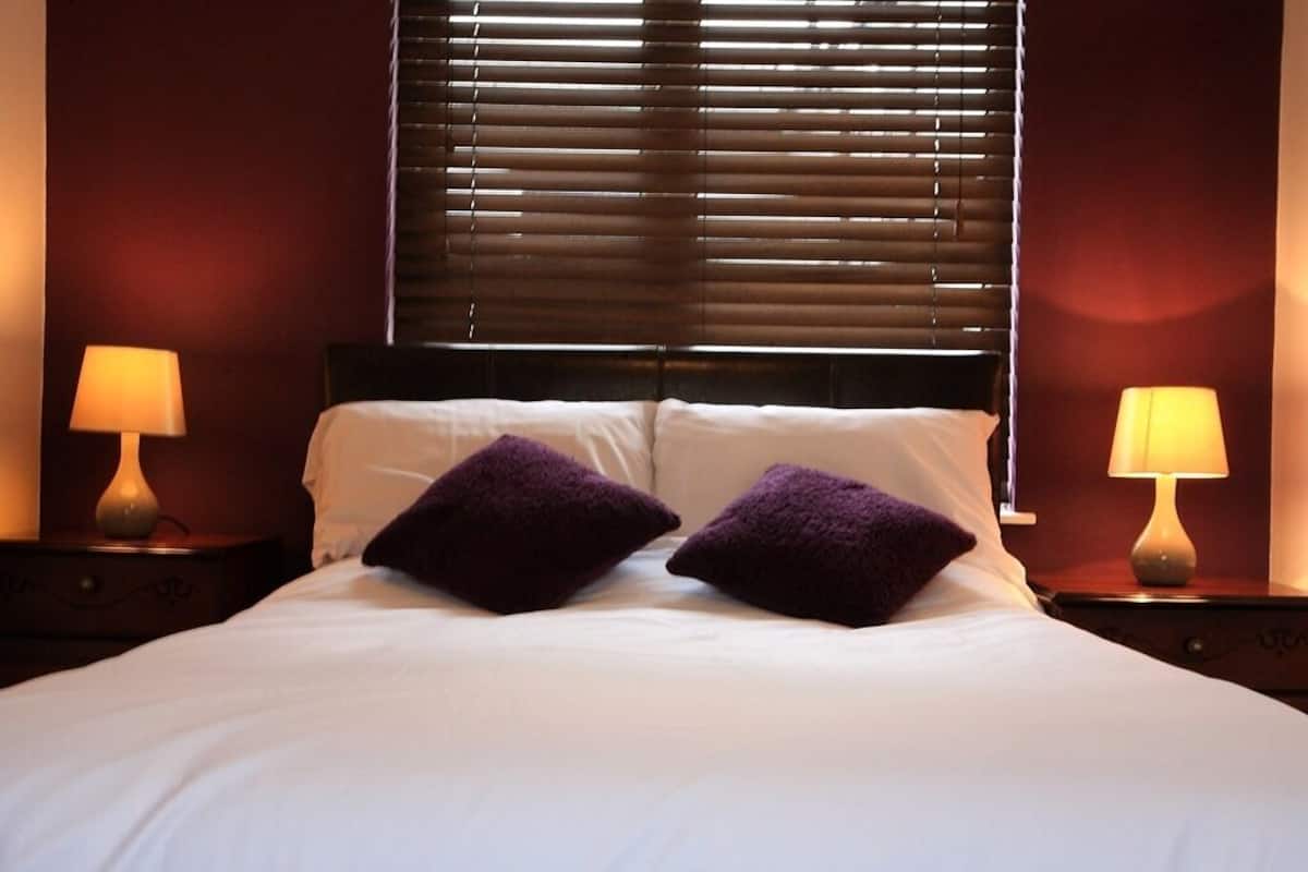 Quarto de hotel com uma cama grande, duas mesas de cabeceira, uma janela com persianas e duas luminárias.