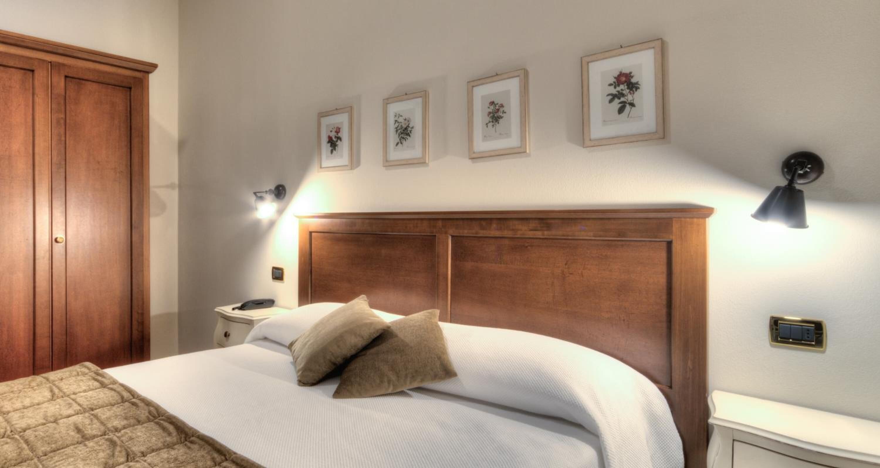 Quarto de hotel com cabeceira de madeira, cama com roupa de cama branca, colcha marrom e quatro gravuras botânicas emolduradas na parede.