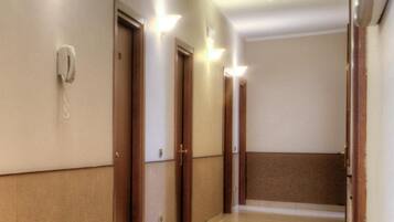 Um corredor comprido e estreito com várias portas, cada uma com uma luz acima. Sinal de saída de emergência verde na parede à direita.
