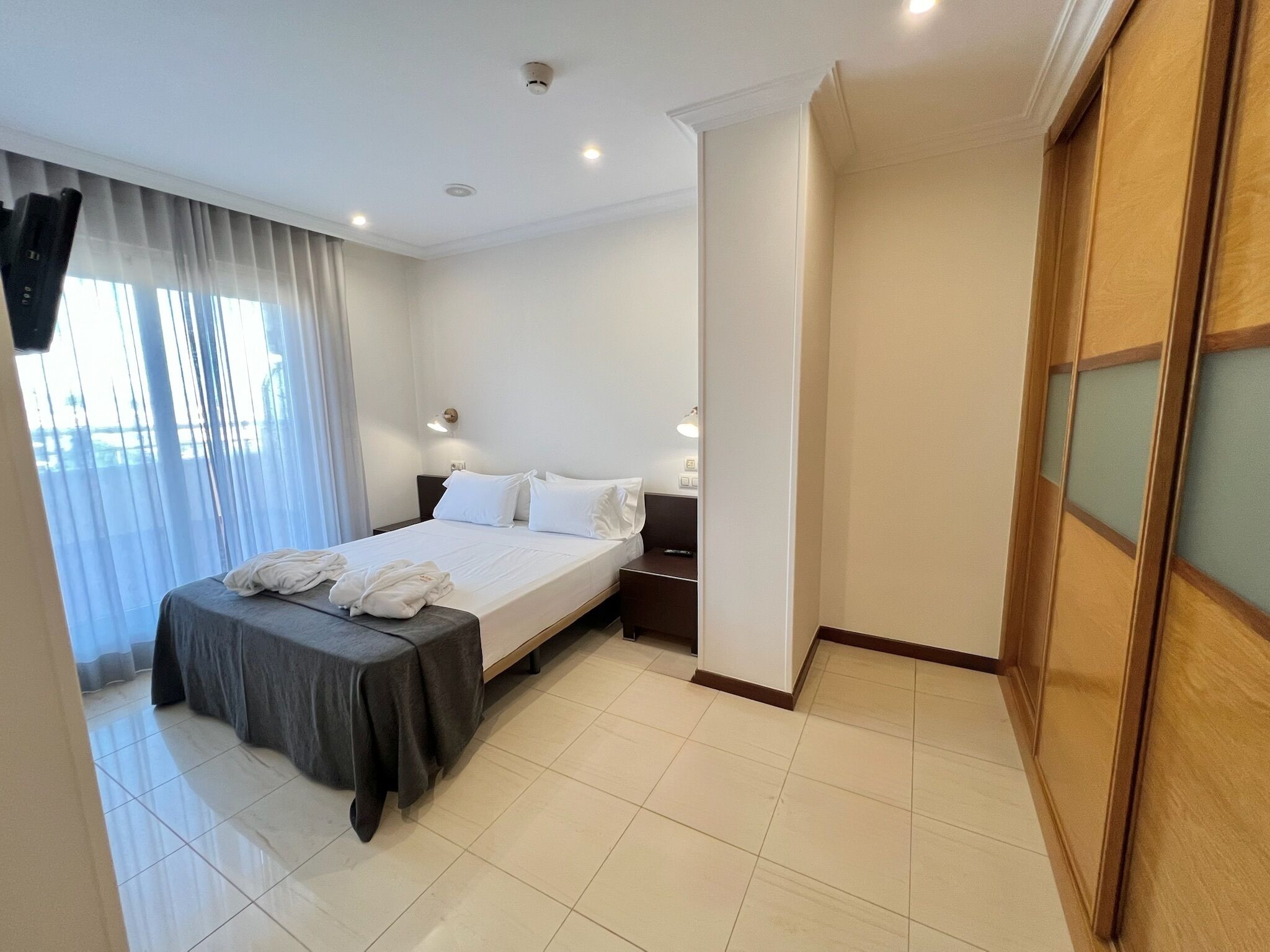 Quarto de hotel com cama, mesas de cabeceira, guarda-roupa e janela com cortinas.