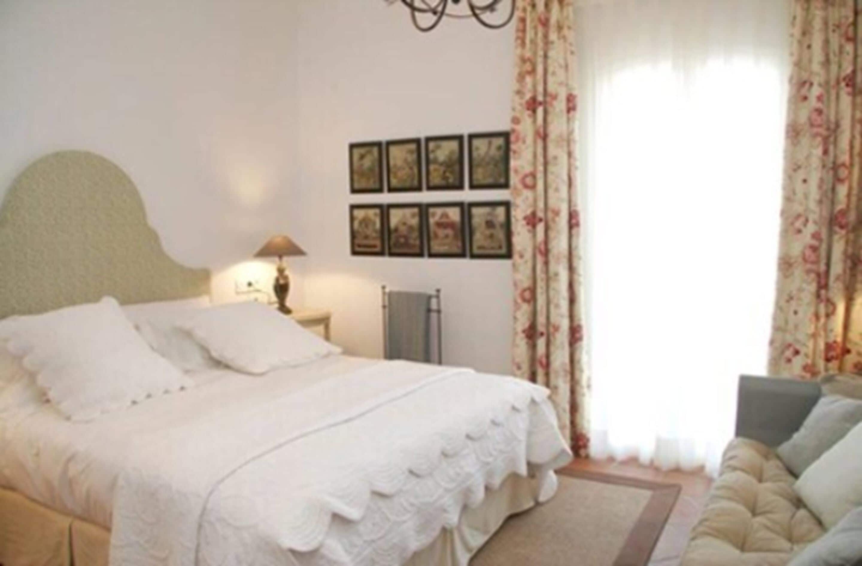 Quarto com cama, mesa de cabeceira, luminária, radiador, sofá e janela com cortinas.