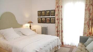 Quarto com cama, mesa de cabeceira, luminária, radiador, sofá e janela com cortinas.
