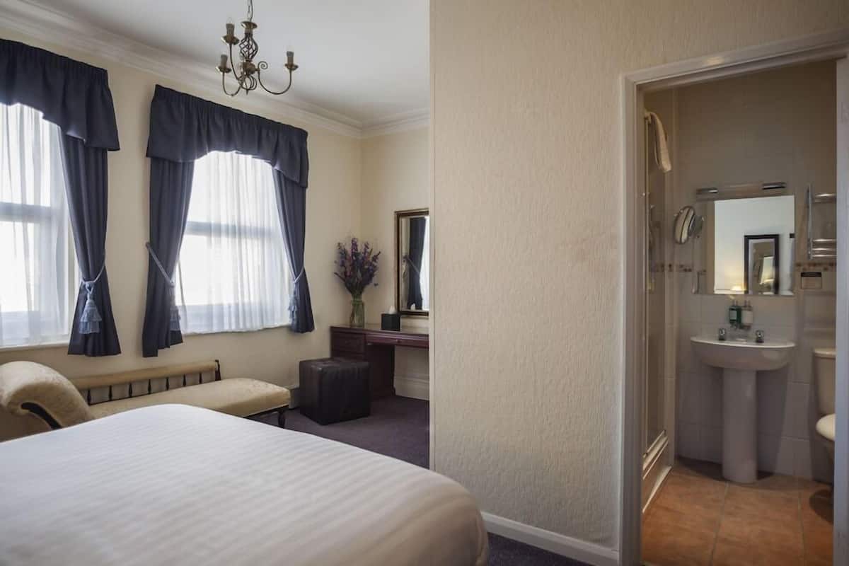Quarto de hotel com cama, um banquinho, escrivaninha e banheiro com pia e espelho.