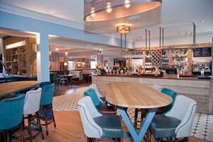 Uma área de bar com mesa de madeira e cadeiras azul-turquesa, balcão bar com diversos frascos e piso estampado.