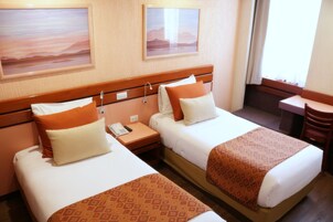 Quarto de hotel com duas camas, cada uma com um edredom estampado, uma mesa de cabeceira com telefone e uma janela com cortinas.