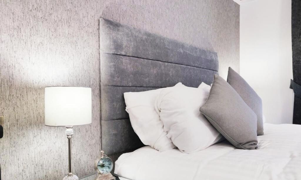 Cama com cabeceira estofada cinza, roupa de cama branca e mesa de cabeceira com abajur e relógio.