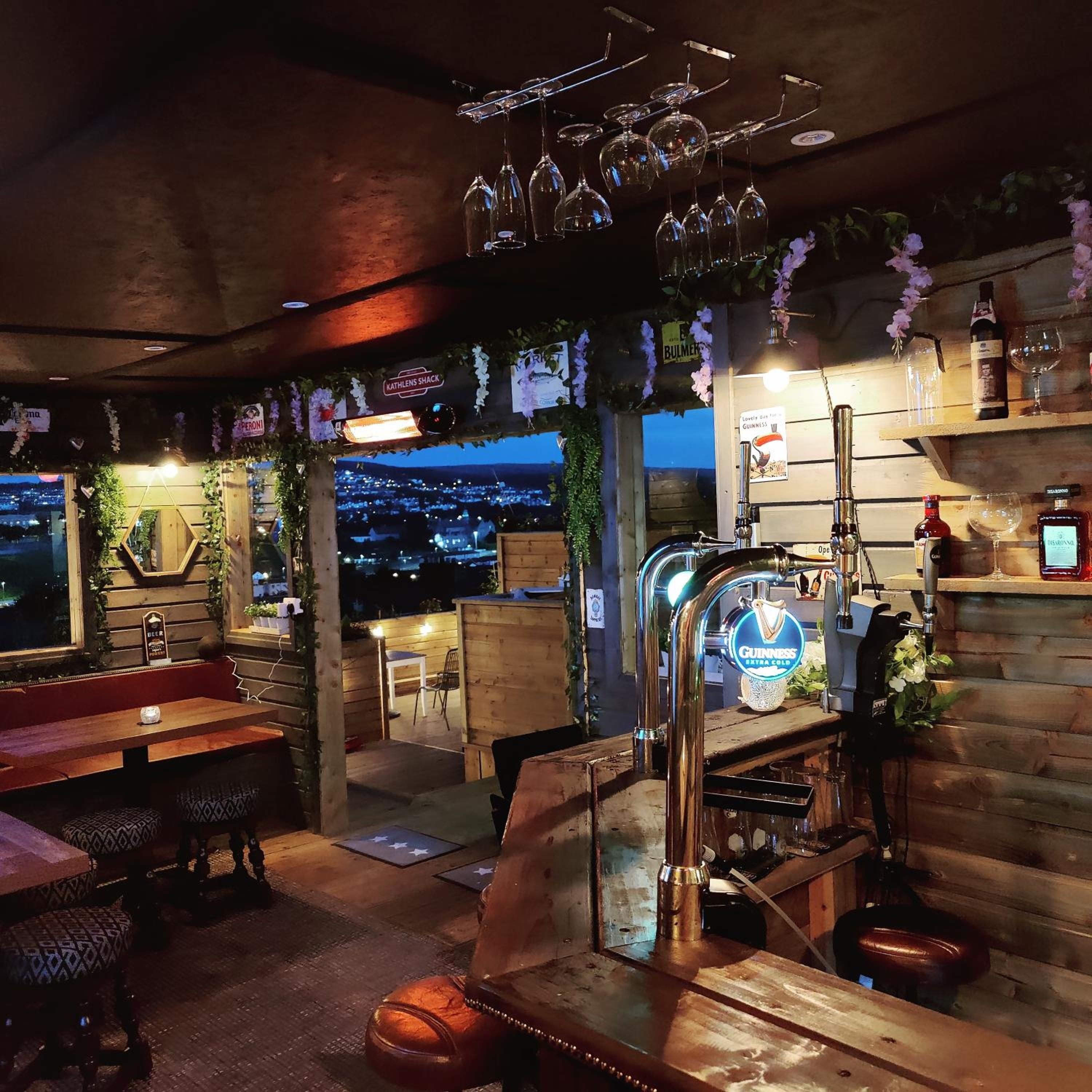 Um espaço de bar acolhedor com mobiliário de madeira, torneira de Guinness e vista para a cidade ao entardecer.