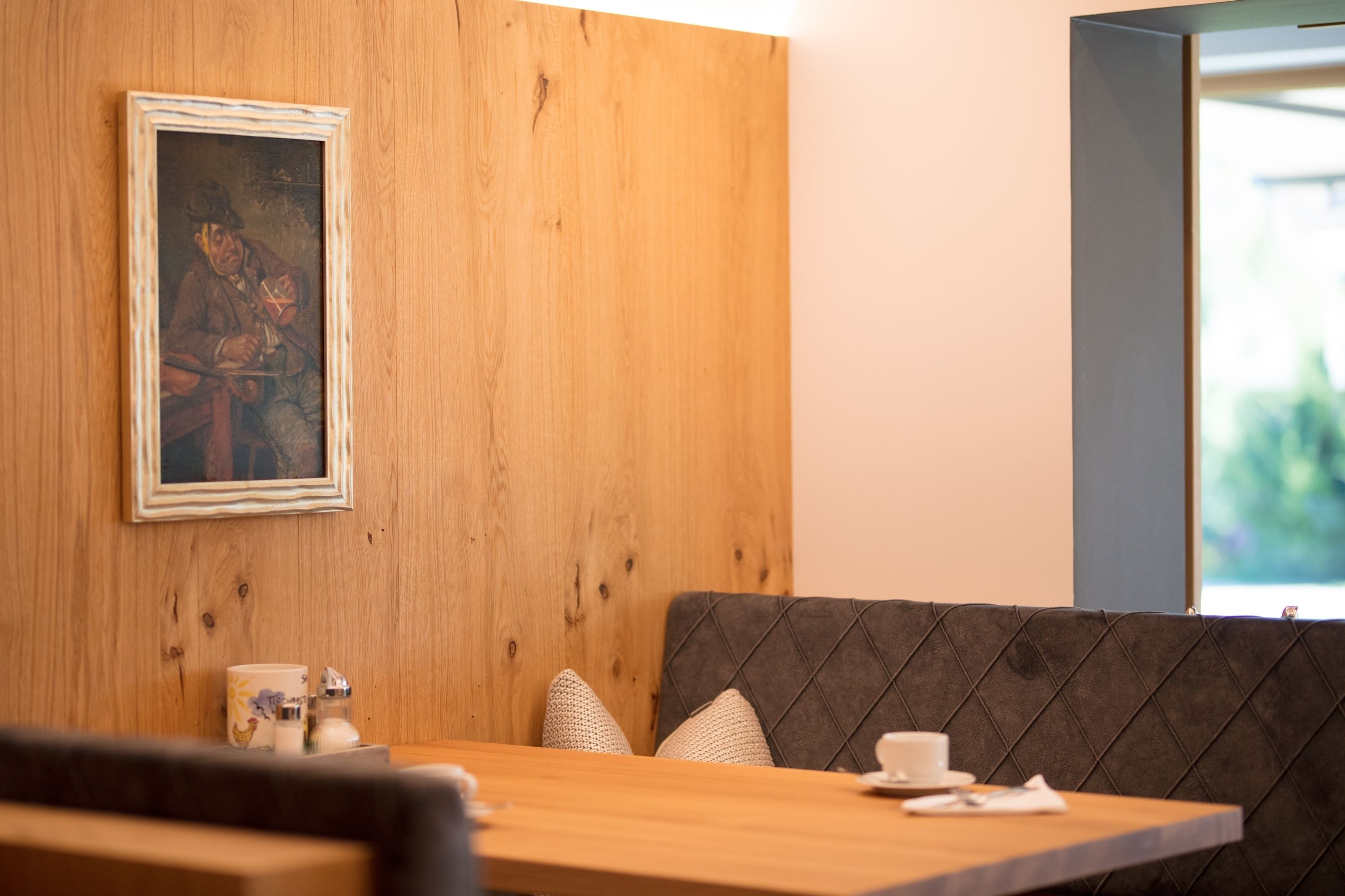 Pared de madeira com um quadro emoldurado, um banco acolchoado, uma mesa com xícara e pires, e uma janela com vista.