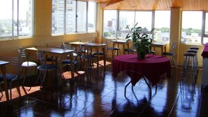 Uma sala de jantar com teto de madeira e janelas amplas, mesas com cadeiras e uma planta em vaso sobre uma das mesas.
