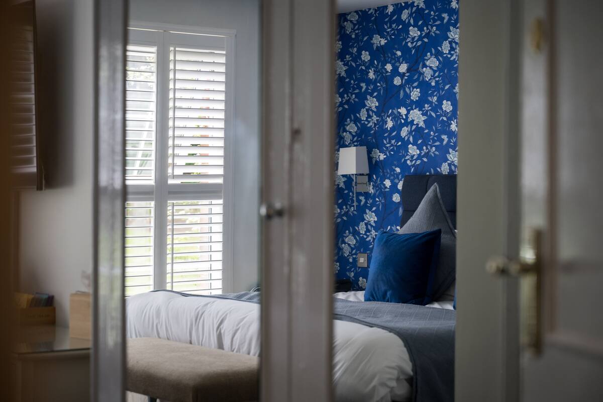 Quarto com cama, mesa de cabeceira, luminária e janela com persianas.