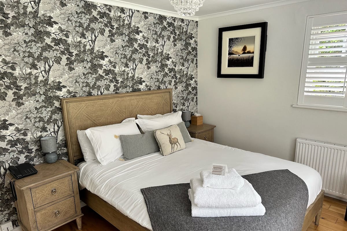 Cama bem arrumada com manta cinza e toalhas brancas, cabeceira de madeira e papel de parede floral.