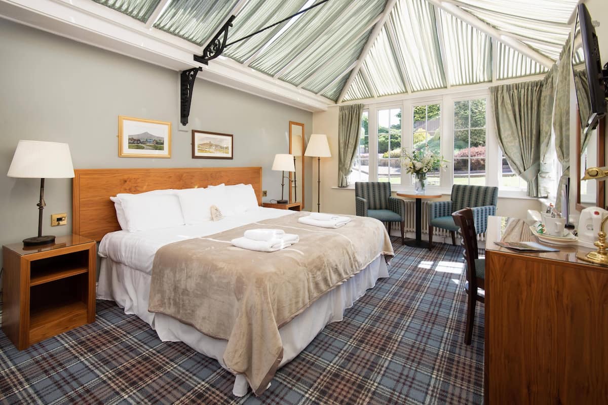 Quarto de hotel com uma cama grande, duas mesas de cabeceira, uma escrivaninha com cadeira, uma pequena mesa com cadeiras e um janelão com cortinas.