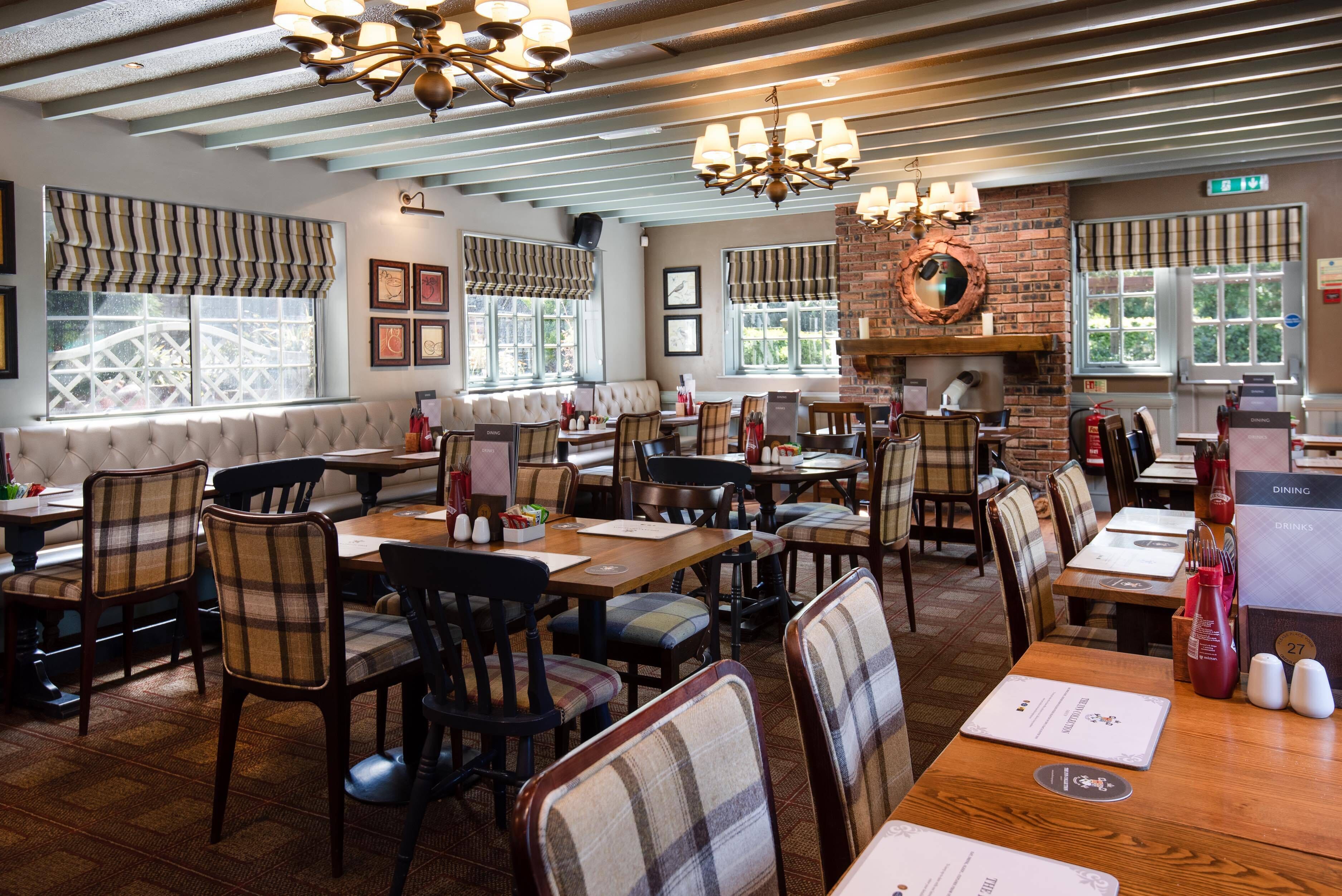 Um restaurante com mesas e cadeiras de madeira, lareira de tijolos e janelas amplas com cortinas.