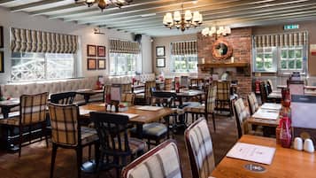 Um restaurante com mesas e cadeiras de madeira, lareira de tijolos e janelas amplas com cortinas.