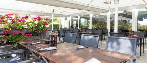 Restaurante ao ar livre com mesas e cadeiras sob um toldo branco, rodeado por vasos de plantas e flores.
