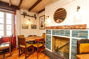 Uma área de refeições com mesa e cadeiras de madeira, lareira com revestimento de azulejos e prateleiras com objetos decorativos.