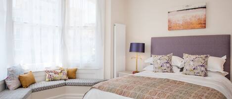 Quarto com cama grande, mesa de cabeceira, abajur e um assento junto à janela com almofadas.