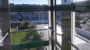 Visão do interior de um elevador, com uma vista clara de um edifício com fachada de vidro extensa e um parque de estacionamento abaixo.